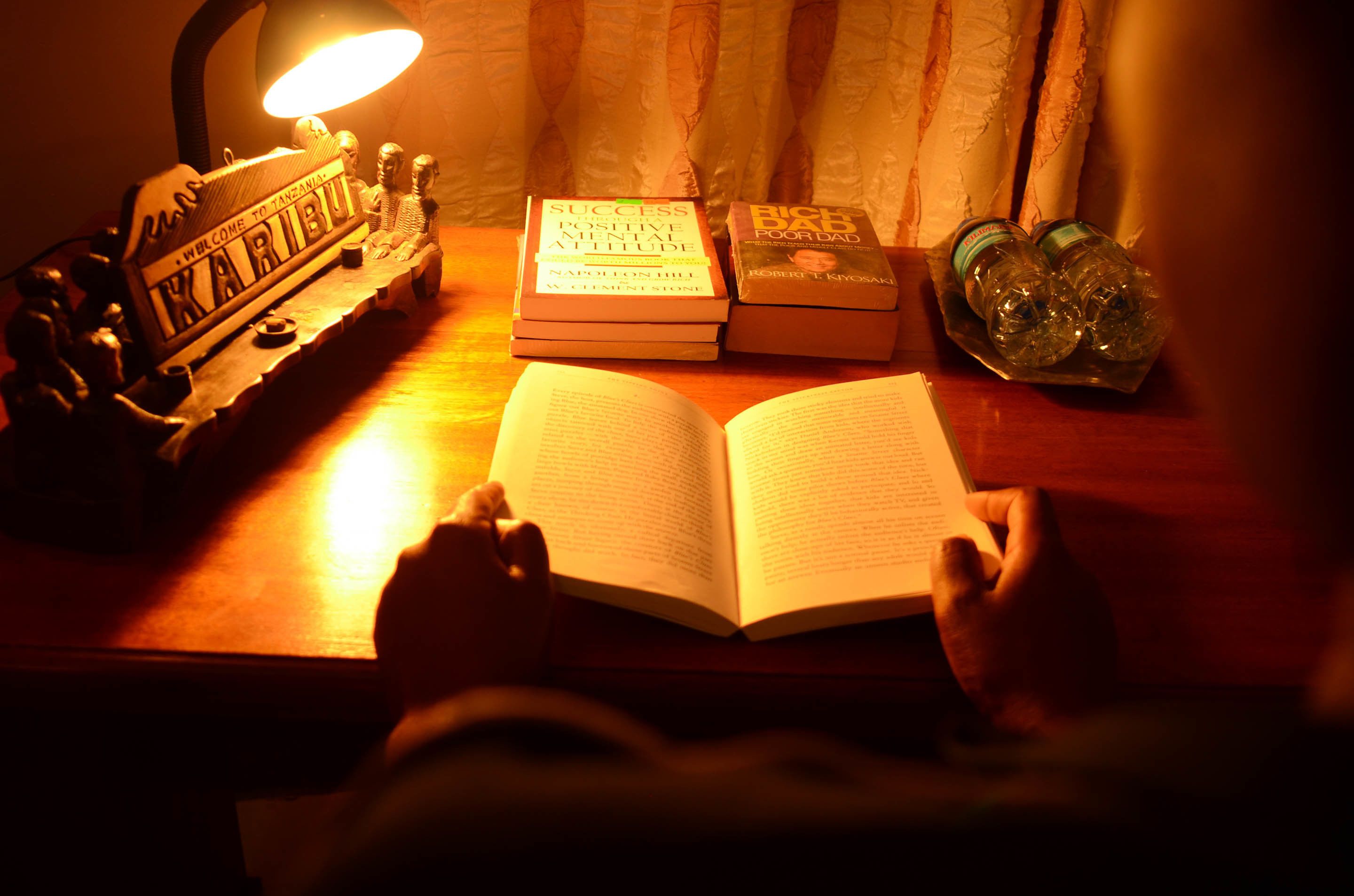 Uma pessoa lendo um livro em uma escrivaninha de madeira com uma luminária, dois livros intitulados 'Success Mindset' e 'Poor Dad', e uma placa com a inscrição 'Welcome to Tanzania'.