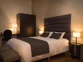 Cama bem arrumada com cabeceira estampada, duas luminárias de apoio, guarda-roupa escuro e um banco listrado.