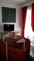 Quarto com televisão sobre uma mesa de vidro, um quadro na parede e cortinas vermelhas.