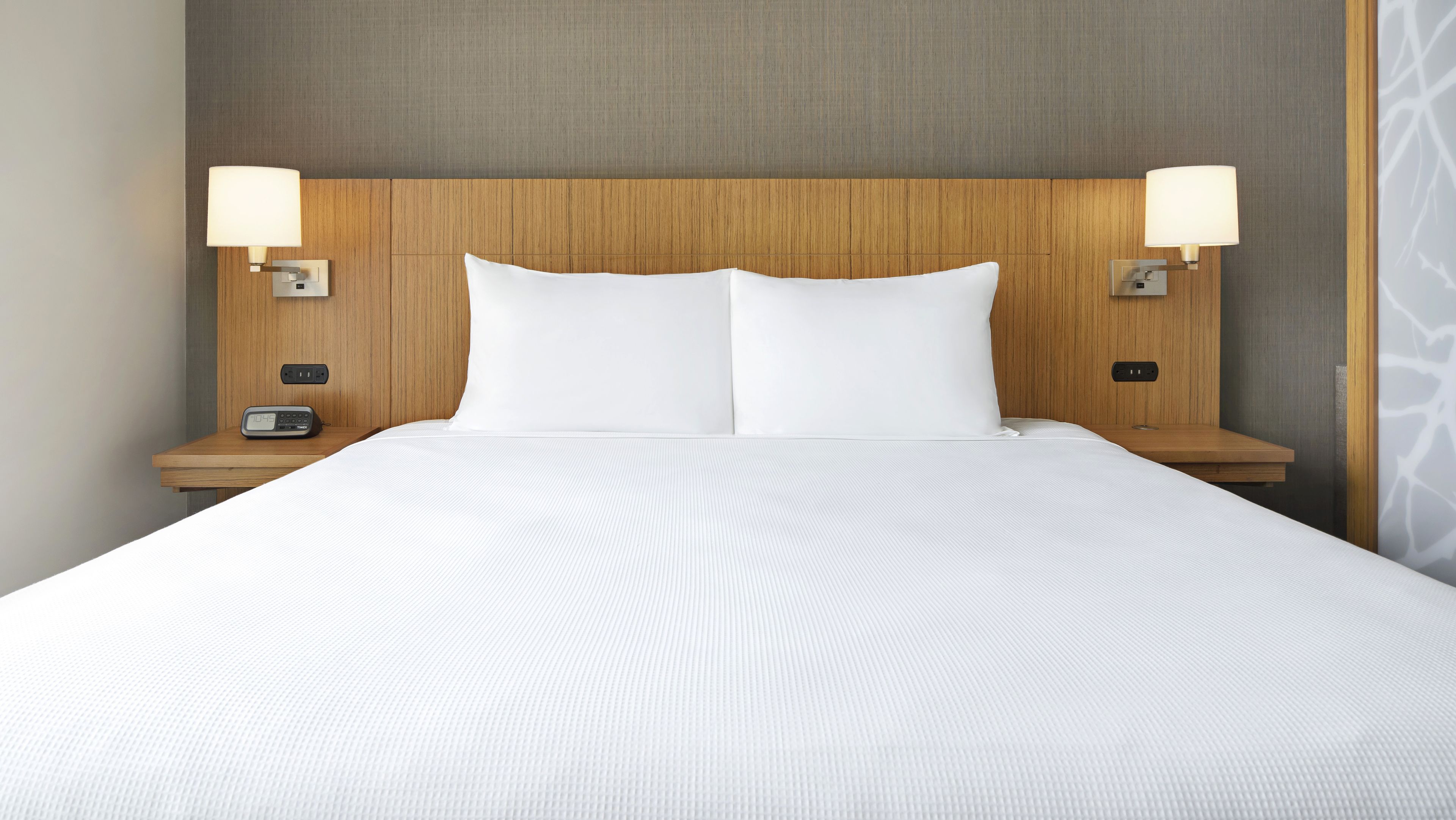 Quarto de hotel com cabeceira de madeira, duas luminárias de apoio, um relógio e uma cama bem arrumada.