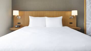 Quarto de hotel com cabeceira de madeira, duas luminárias de apoio, um relógio e uma cama bem arrumada.