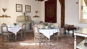 Uma área de jantar com uma mesa redonda posta para quatro pessoas, rodeada por quatro cadeiras. Há um banco de madeira com mesa e uma cadeira no canto. O ambiente possui piso de azulejo e teto de madeira com vigas aparentes.