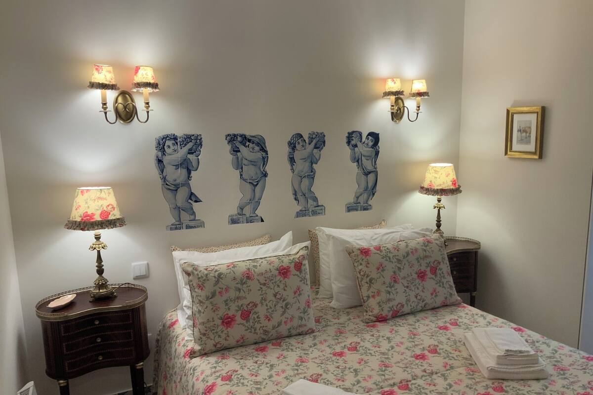 Quarto com roupa de cama florida, duas luminárias de cabeceira, luminária na parede e um quadro emoldurado.