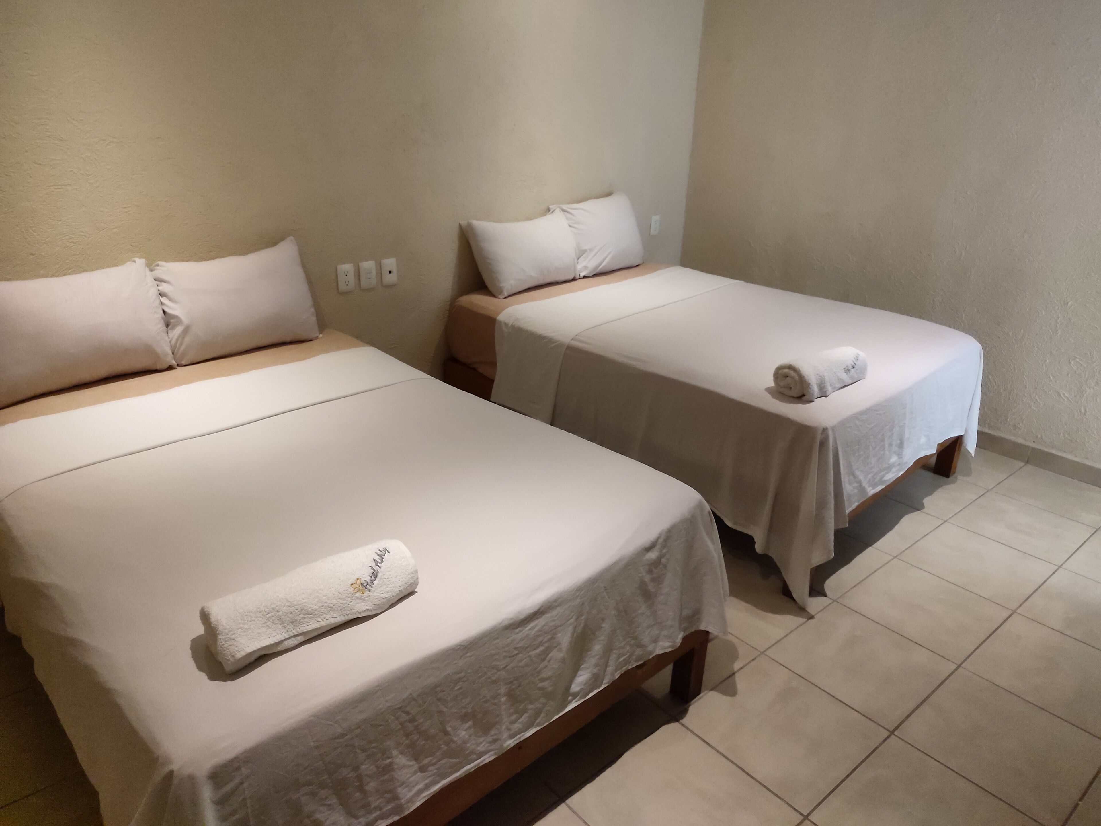 Duas camas de solteiro com lençóis e almofadas brancos, uma pequena mesa de cabeceira e uma tomada na parede.