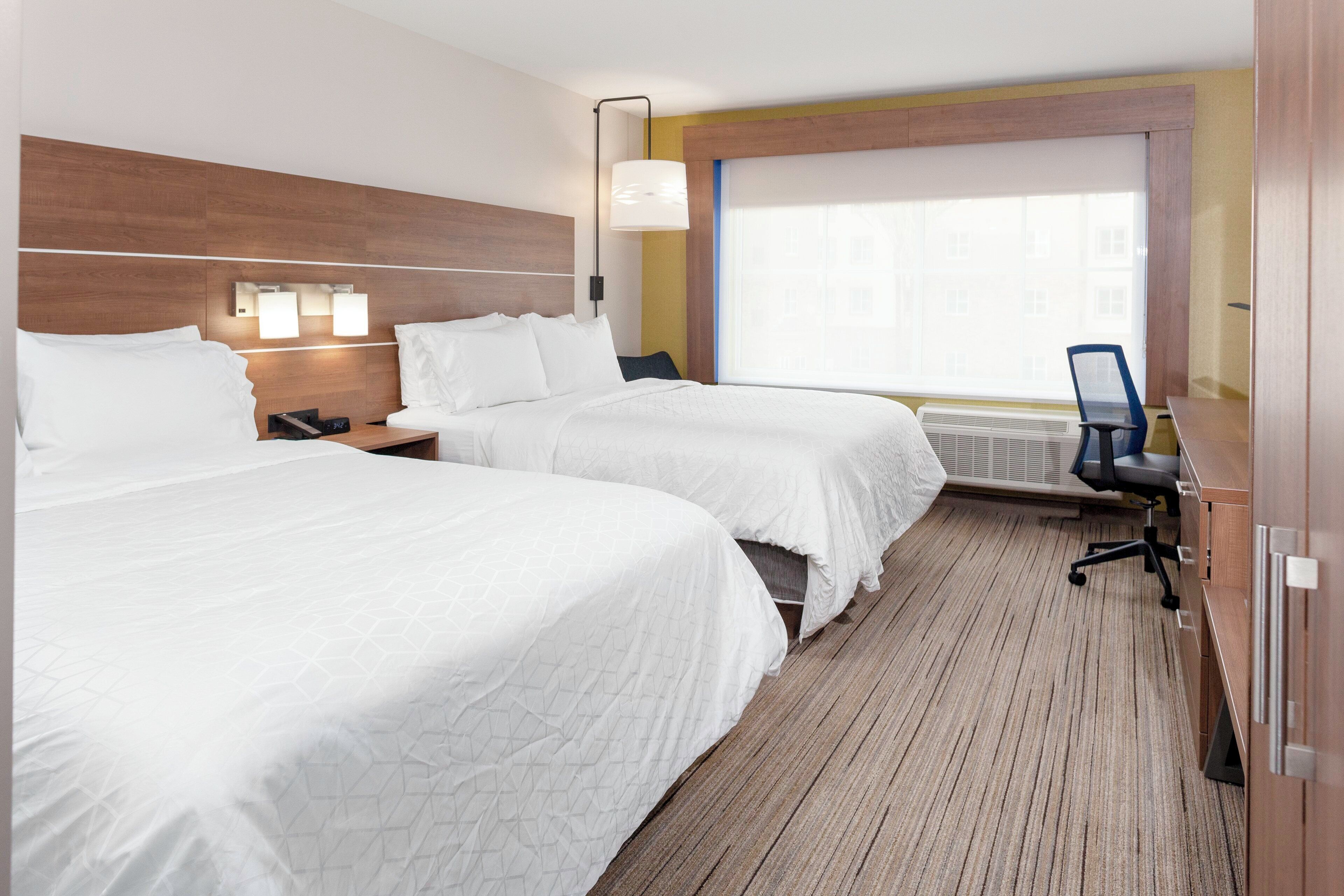 Quarto de hotel com duas camas, uma escrivaninha com cadeira, uma janela com persianas e um abajur fixado na parede.