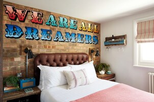 Quarto com parede de tijolos, cabeceira estofada, cama com roupa de cama branca, mesa de cabeceira com abajur e placa na parede com a inscrição 'WE ARE ALL DREAMERS'.