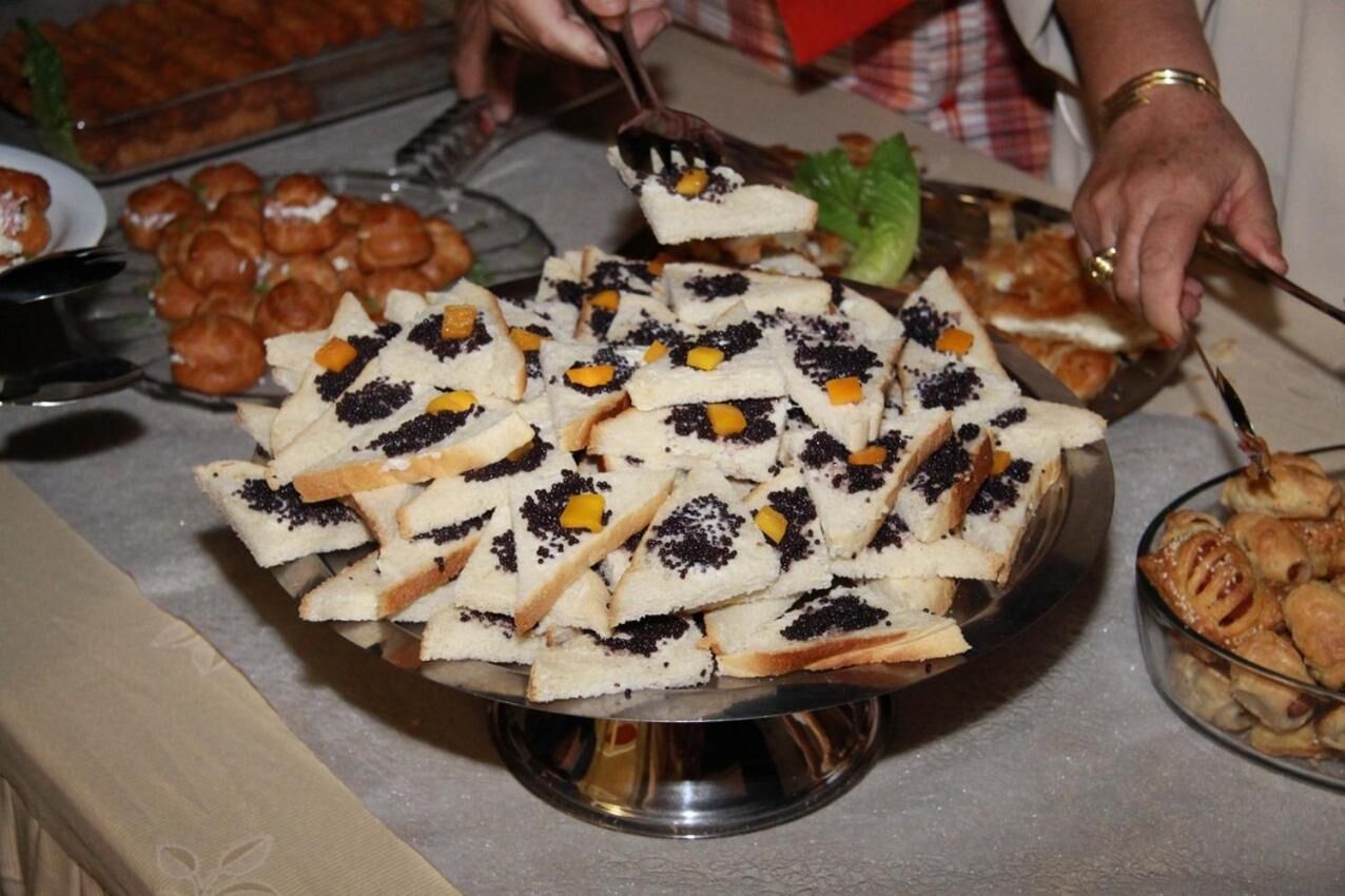 Um buffet com diversos alimentos, incluindo pão com sementes de papoula e pedaços de laranja, e uma pessoa servindo.