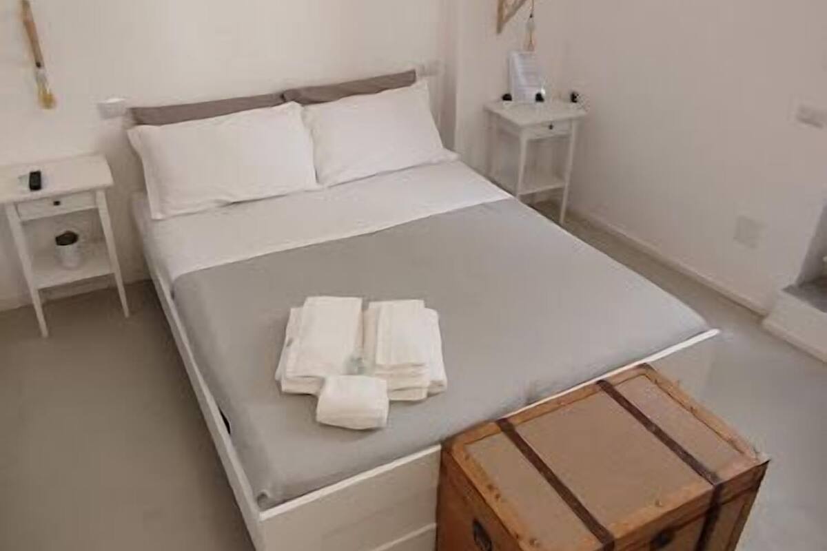 Quarto minimalista com uma cama, mesa de cabeceira e mala.