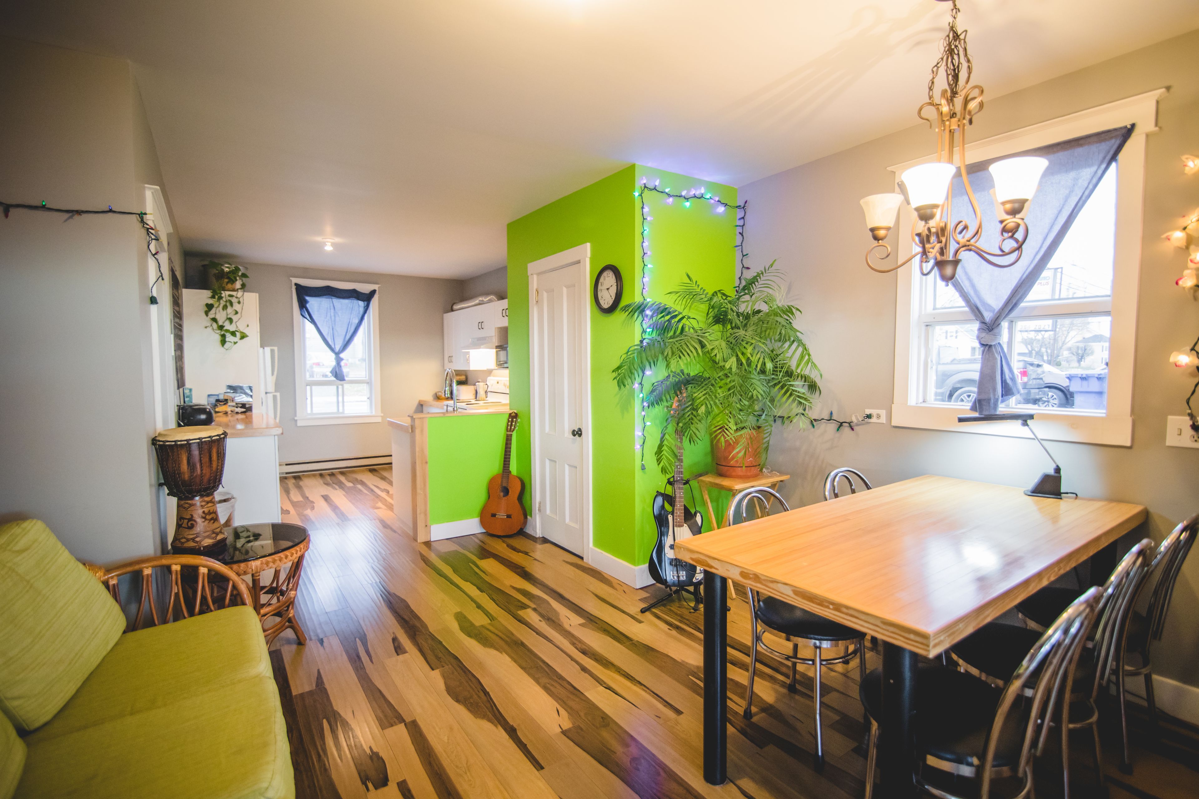 Uma área de refeições com mesa e cadeiras de madeira, uma parede verde, uma planta em vaso e um violão encostado na parede.