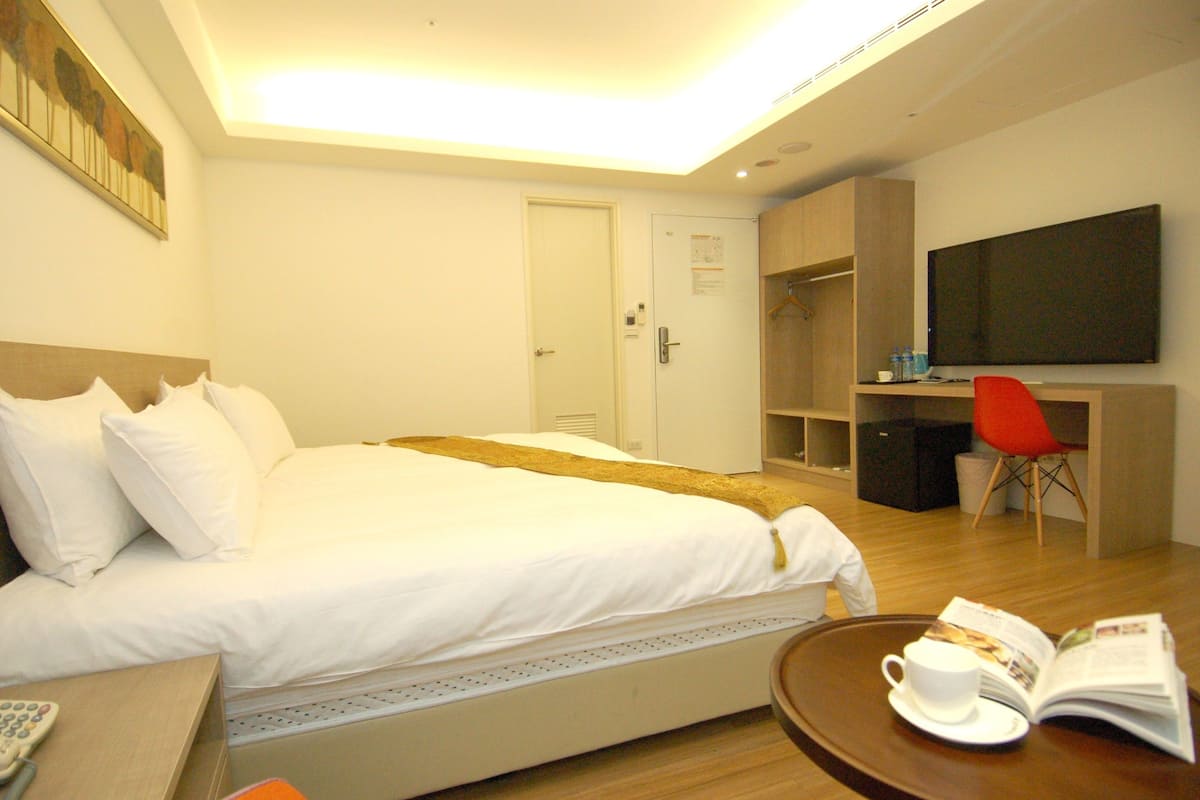 Quarto de hotel com uma cama, uma escrivaninha, uma TV e uma mesa de centro com um livro e uma xícara.