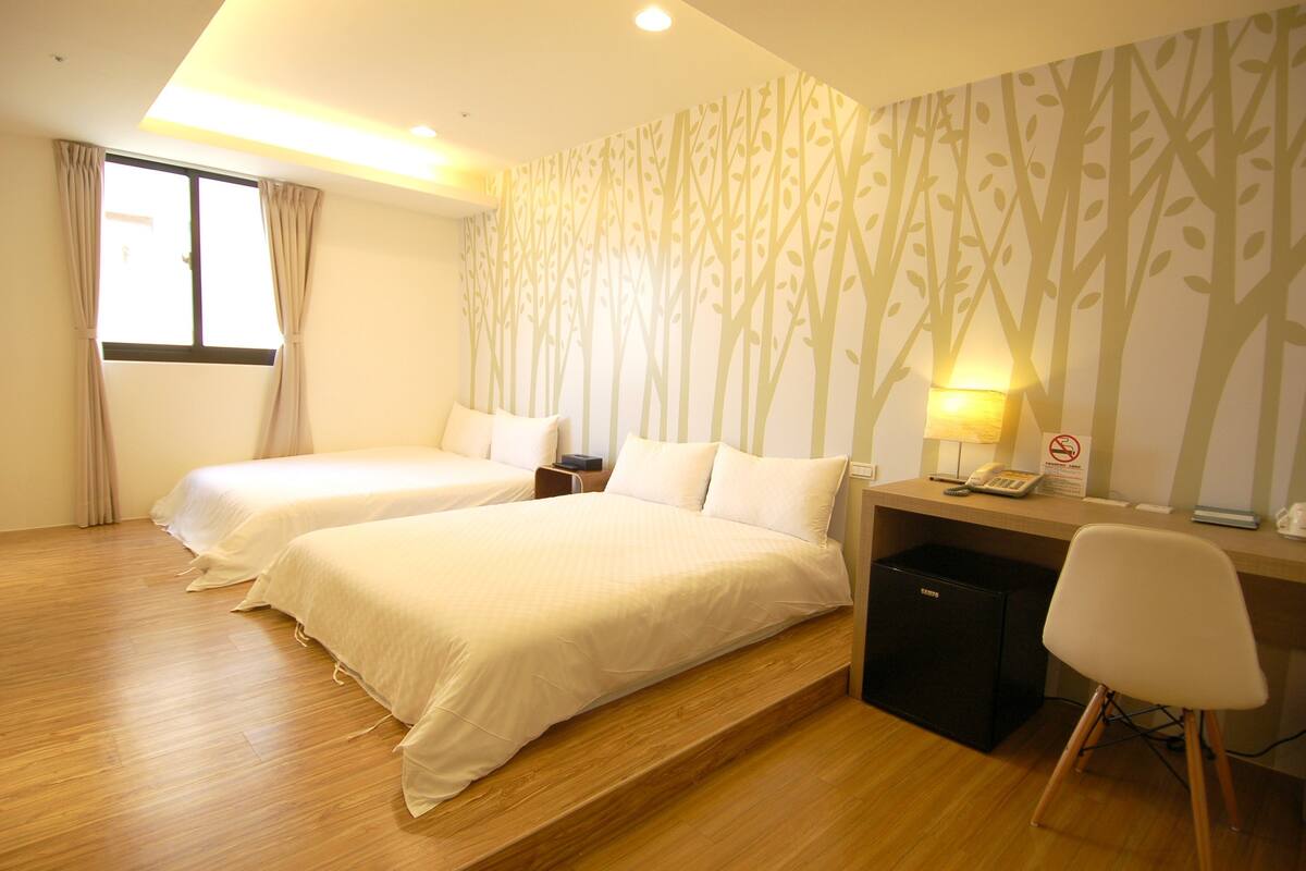 Quarto de hotel com duas camas, uma escrivaninha, uma cadeira, um cesto de lixo e uma janela com cortinas.