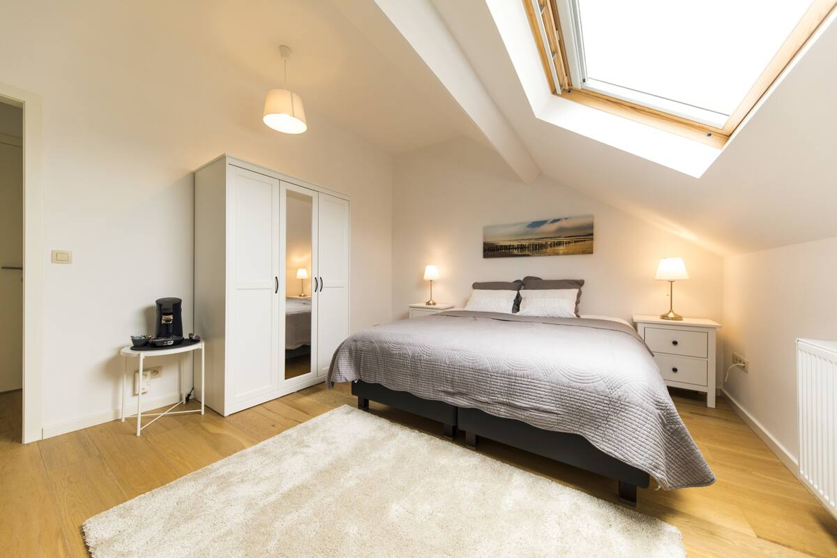 Quarto com claraboia, cama com colcha cinza, mesas de cabeceira com abajures e um guarda-roupa branco.