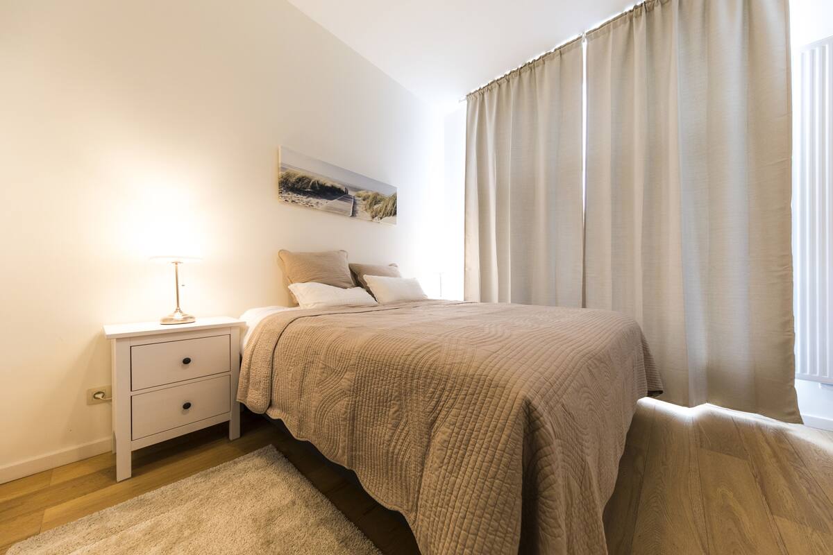 Quarto com cama, mesa de cabeceira, abajur, um quadro na parede e janela com cortinas.