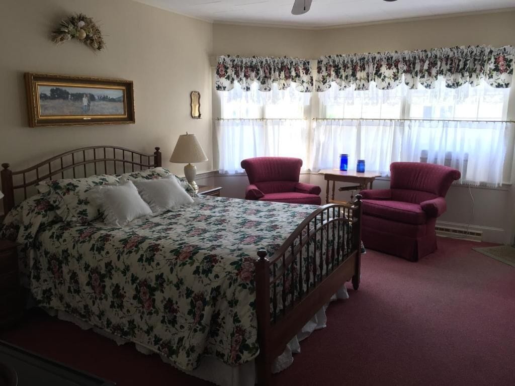 Quarto com cama de madeira, duas poltronas iguais, uma mesinha e janela com cortinas florais.