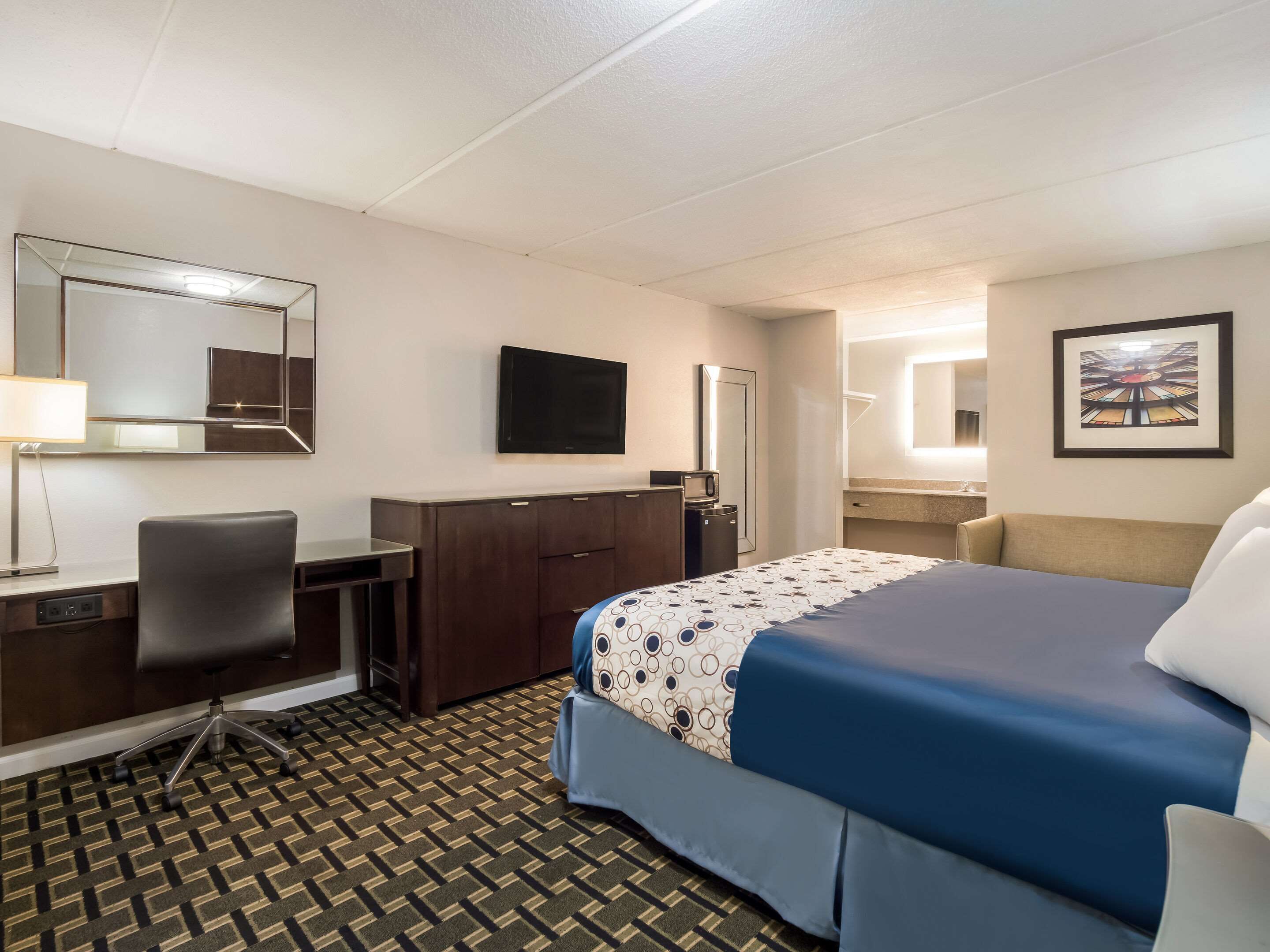 Quarto de hotel com cama, escrivaninha, cadeira, televisão, espelho e quadro na parede.