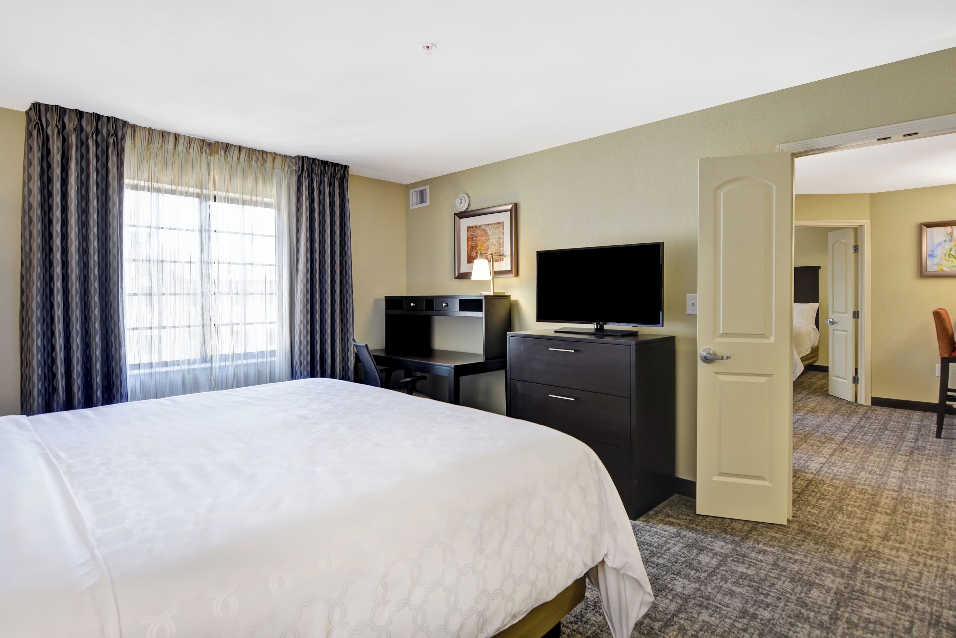 Quarto de hotel com cama, escrivaninha com televisão, cadeira e porta que dá para outro quarto.