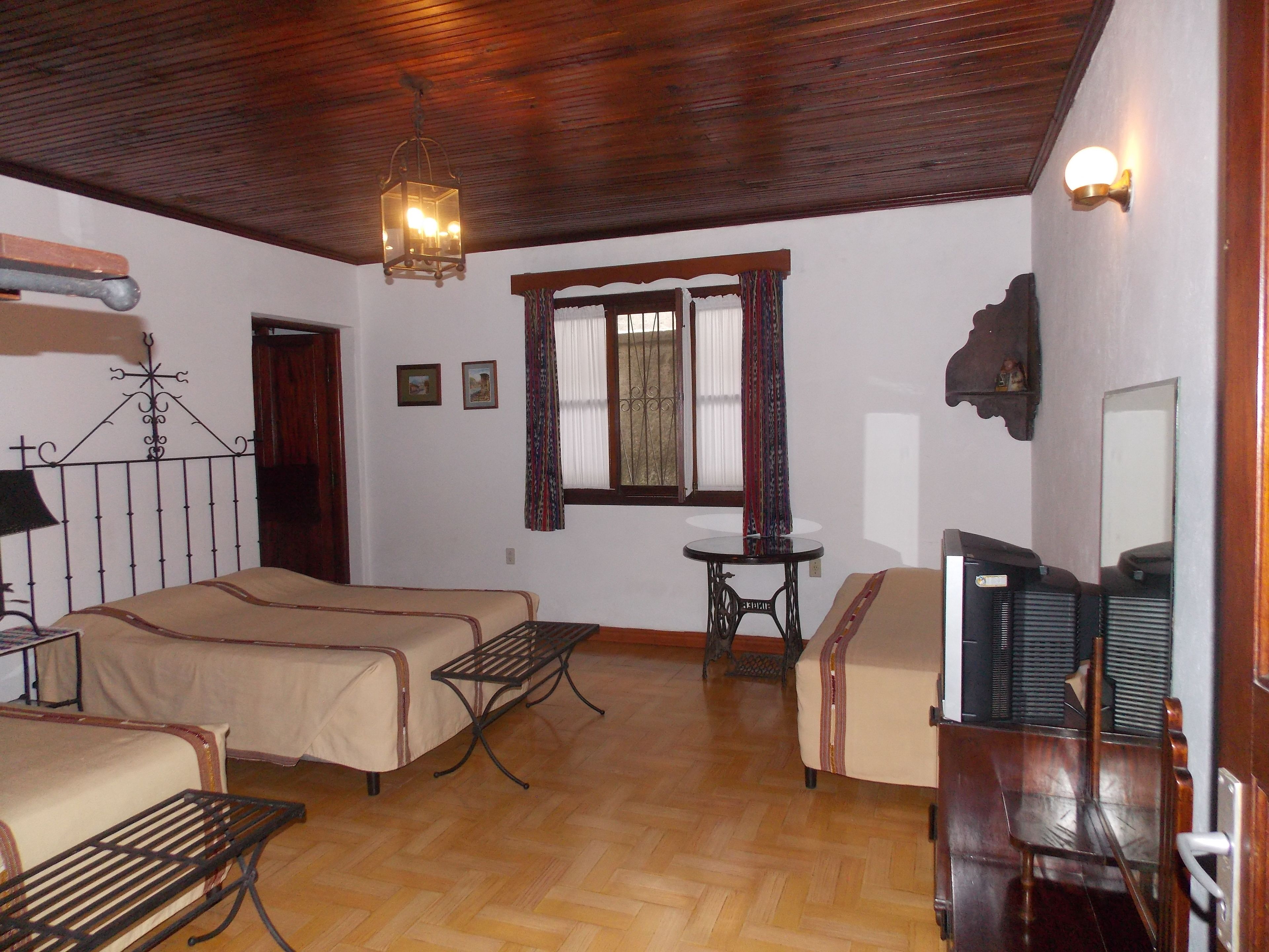 Quarto com duas camas, uma mesinha, uma cadeira, televisão e janela com cortinas.