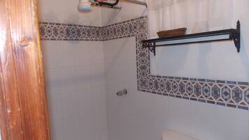 Casa de banho com azulejos brancos, chuveiro, sanita e uma prateleira com uma cesta.