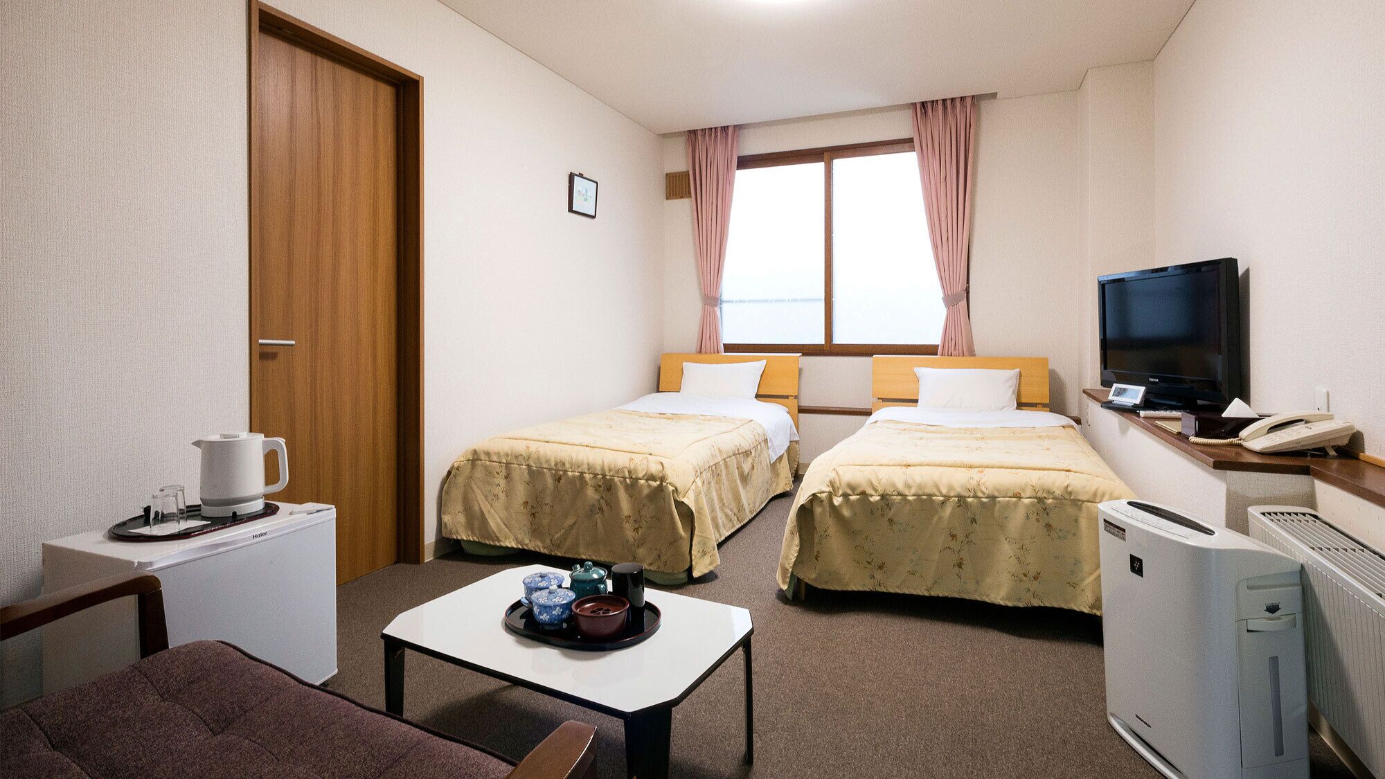 Quarto de hotel com duas camas, televisão, secretária com chaleira, pequena mesa com louças e radiador.