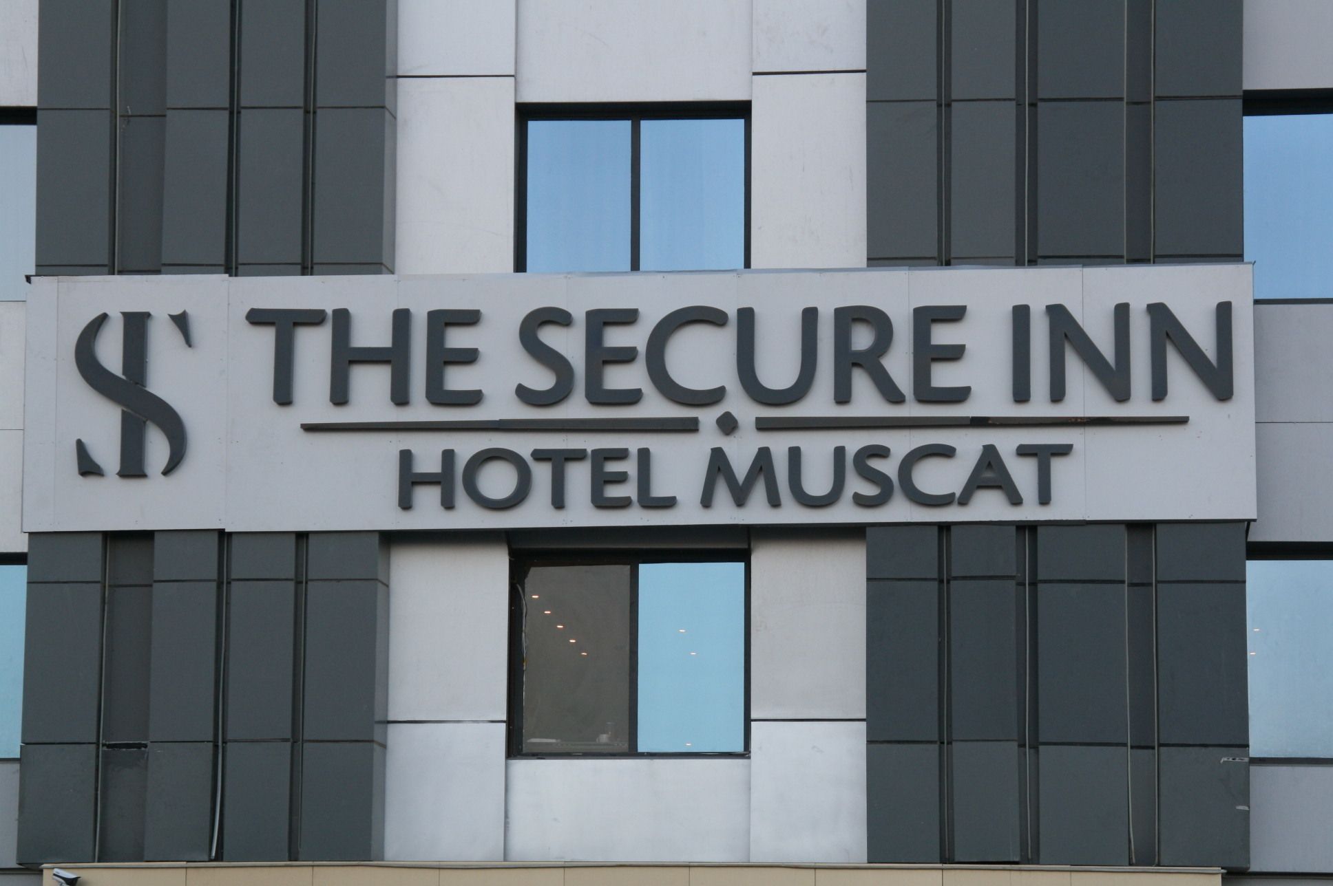 Placa do Hotel The Secure Inn Muscat com design arquitetônico moderno, caracterizado por grandes janelas e fachada geométrica e clean.