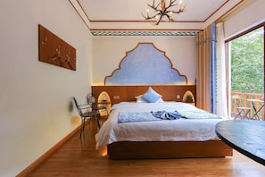 Quarto com cama grande, lustre, cabeceira decorativa, mesa de jantar e vista para o verde lá fora.