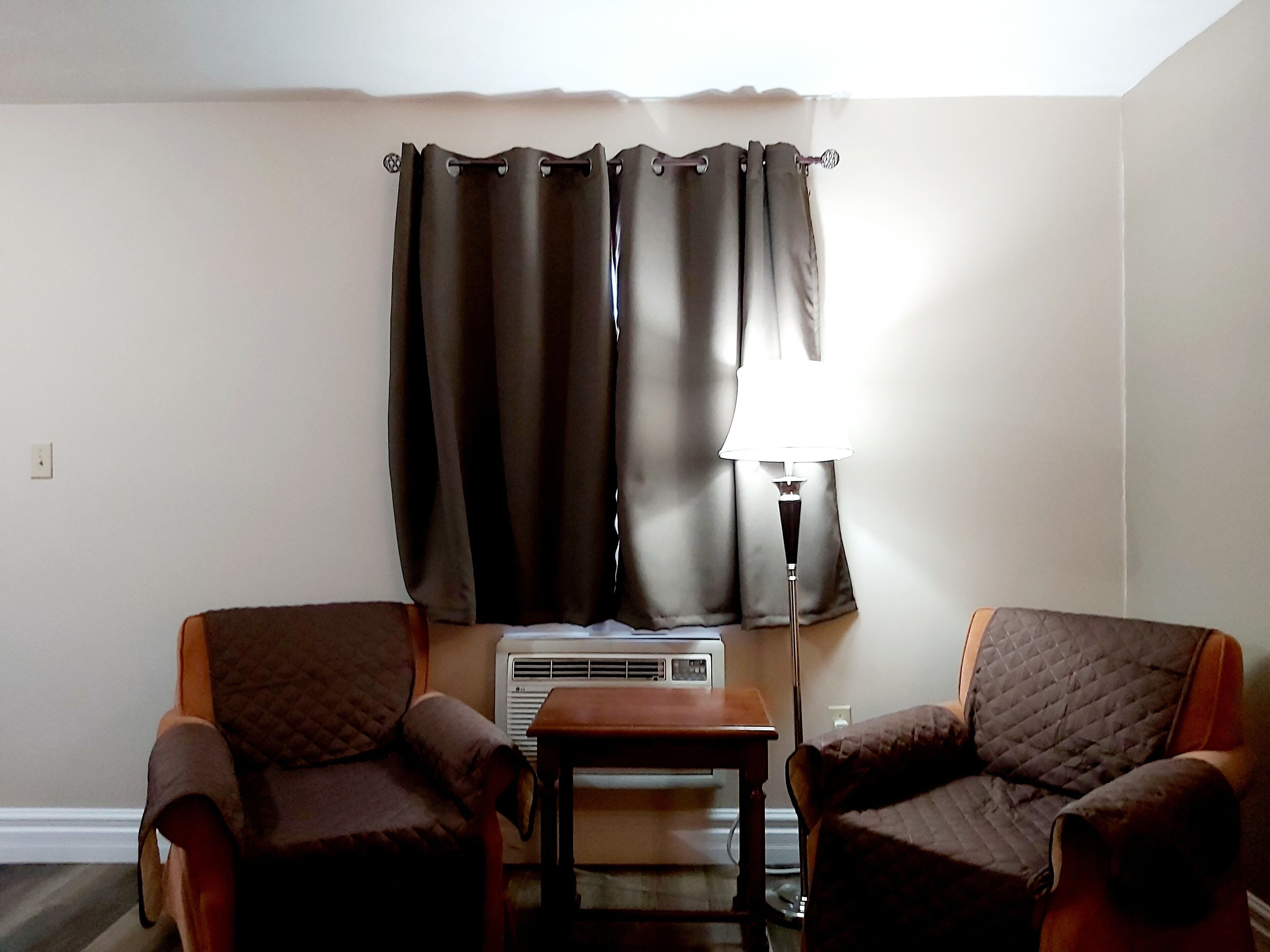 Um quarto com duas poltronas marrons, uma mesinha, uma luminária e um aparelho de ar condicionado.