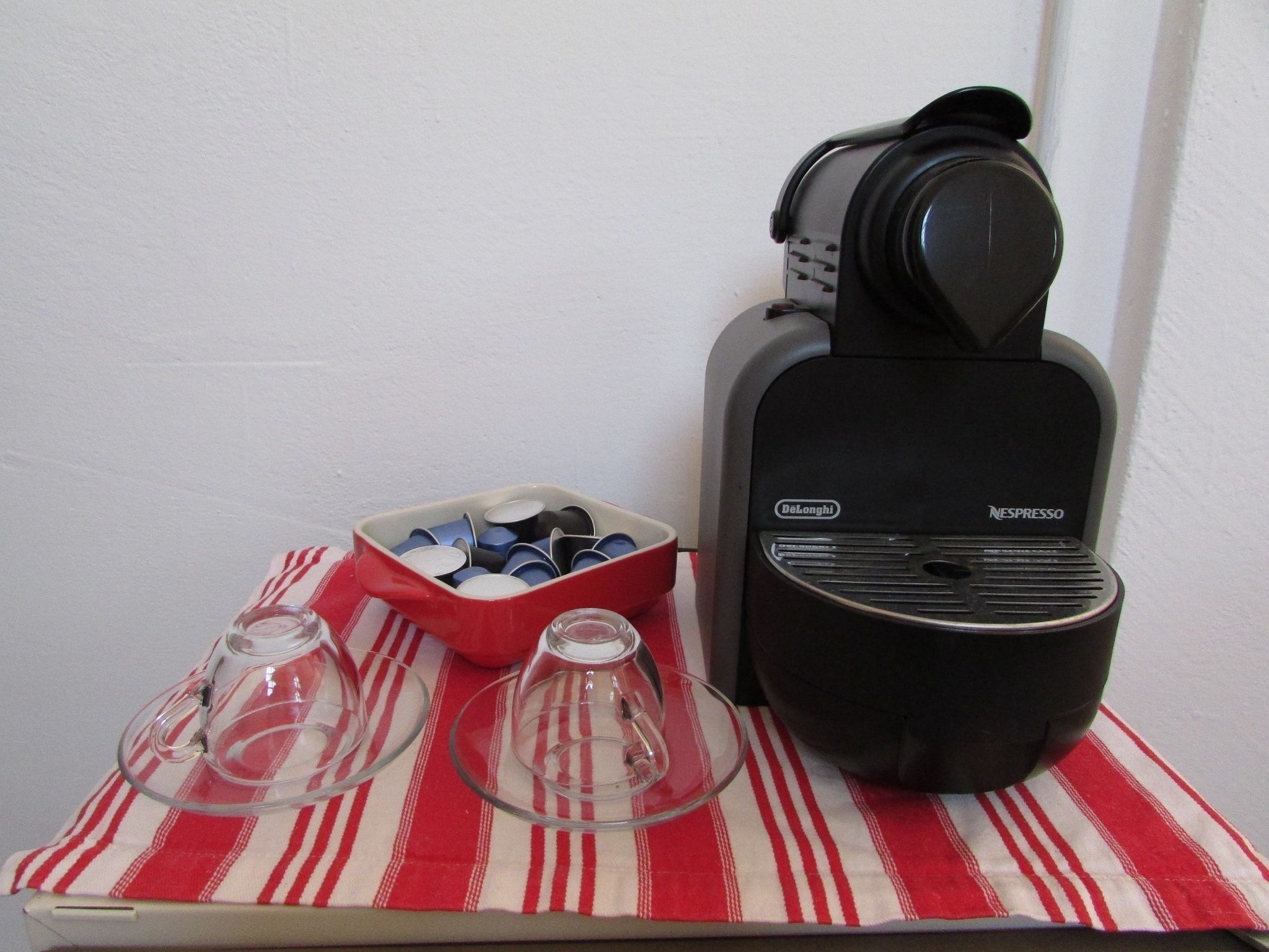 Uma máquina de café DeLonghi Nespresso, um pano listrado em vermelho e branco e uma tigela com cápsulas de café.