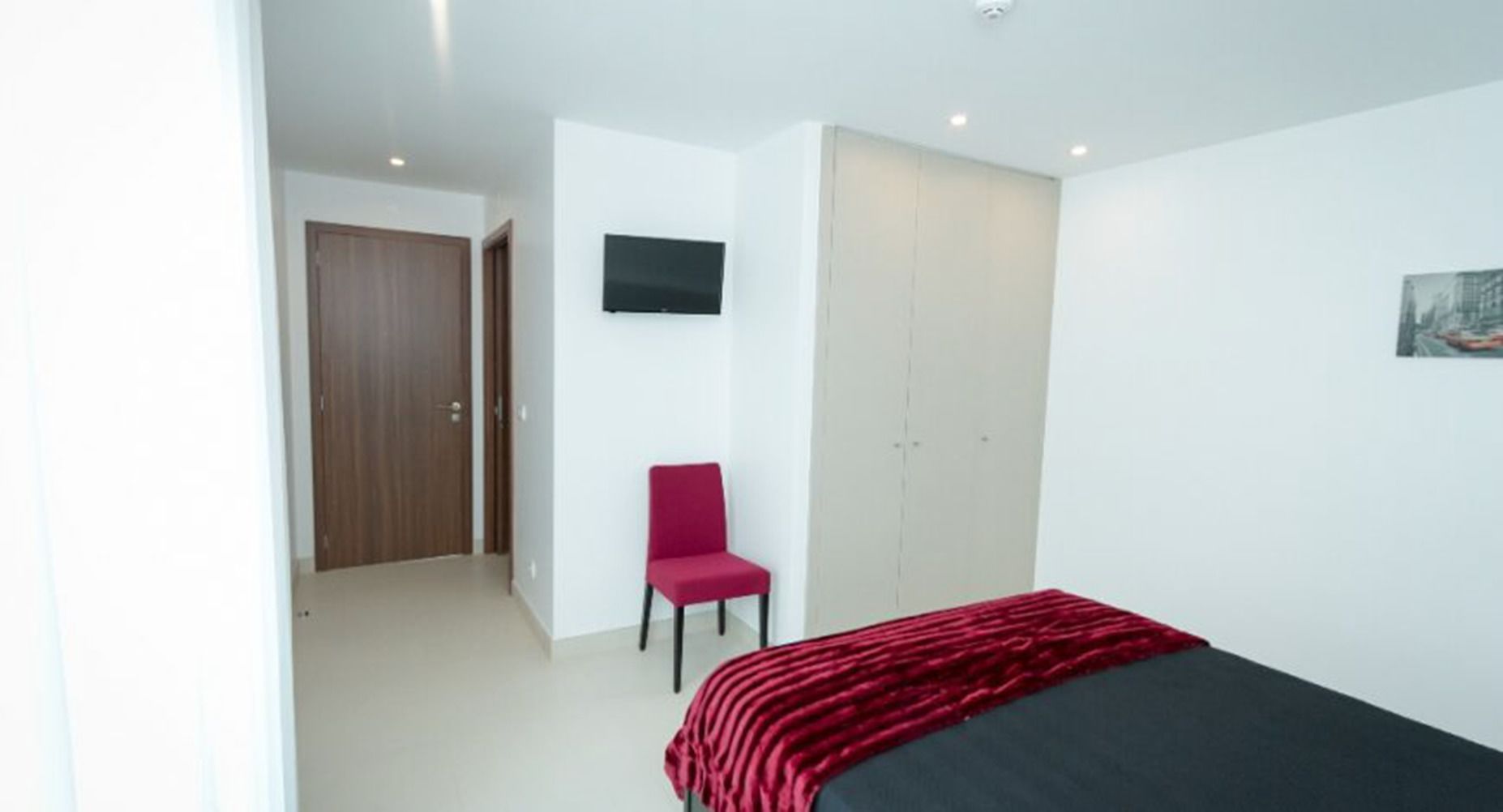 Um quarto com uma cama, uma cadeira vermelha, um guarda-roupa e uma televisão na parede.