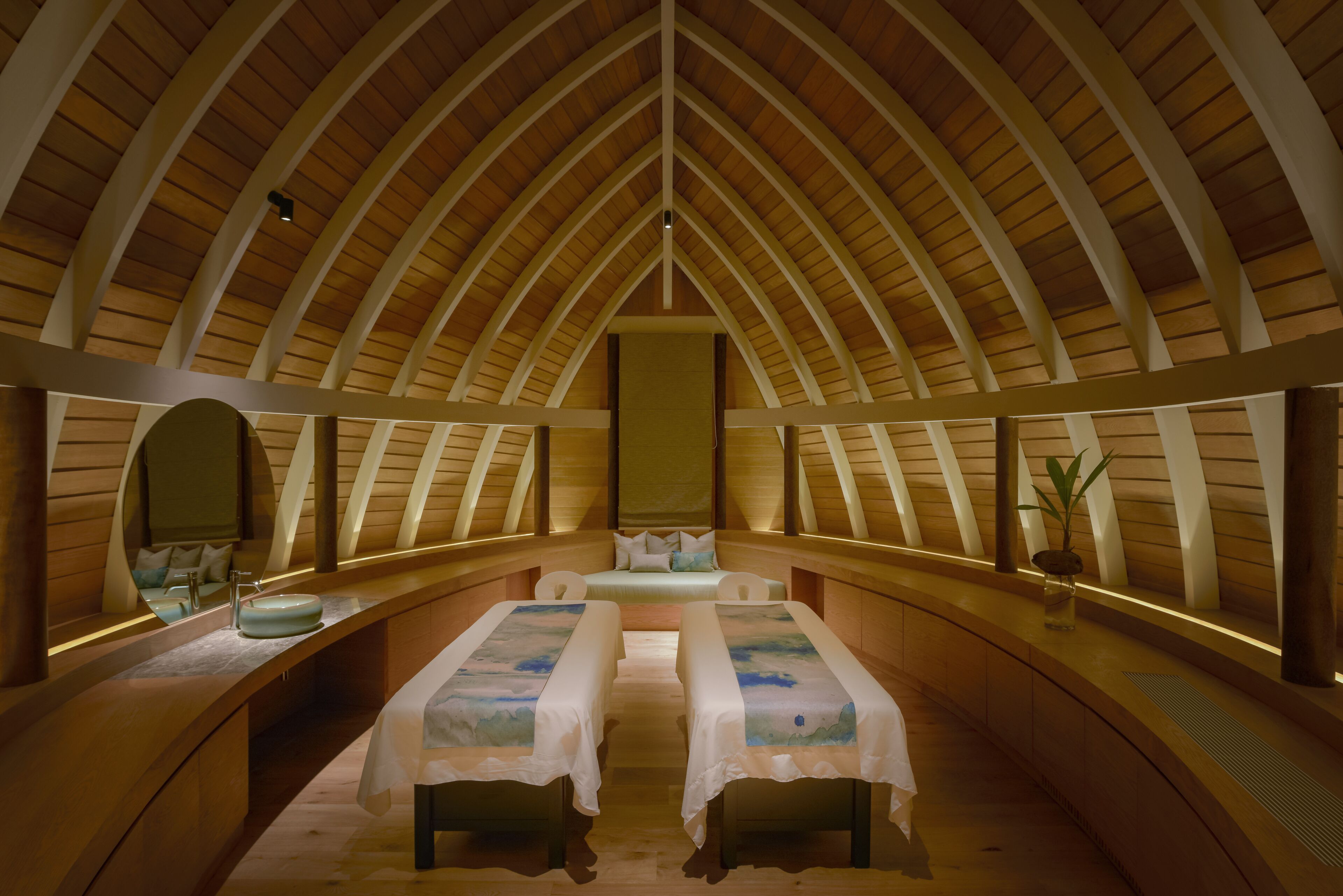 Quarto com teto de madeira abobadado, duas camas com cabeceiras iguais, um espelho grande e uma planta pequena.