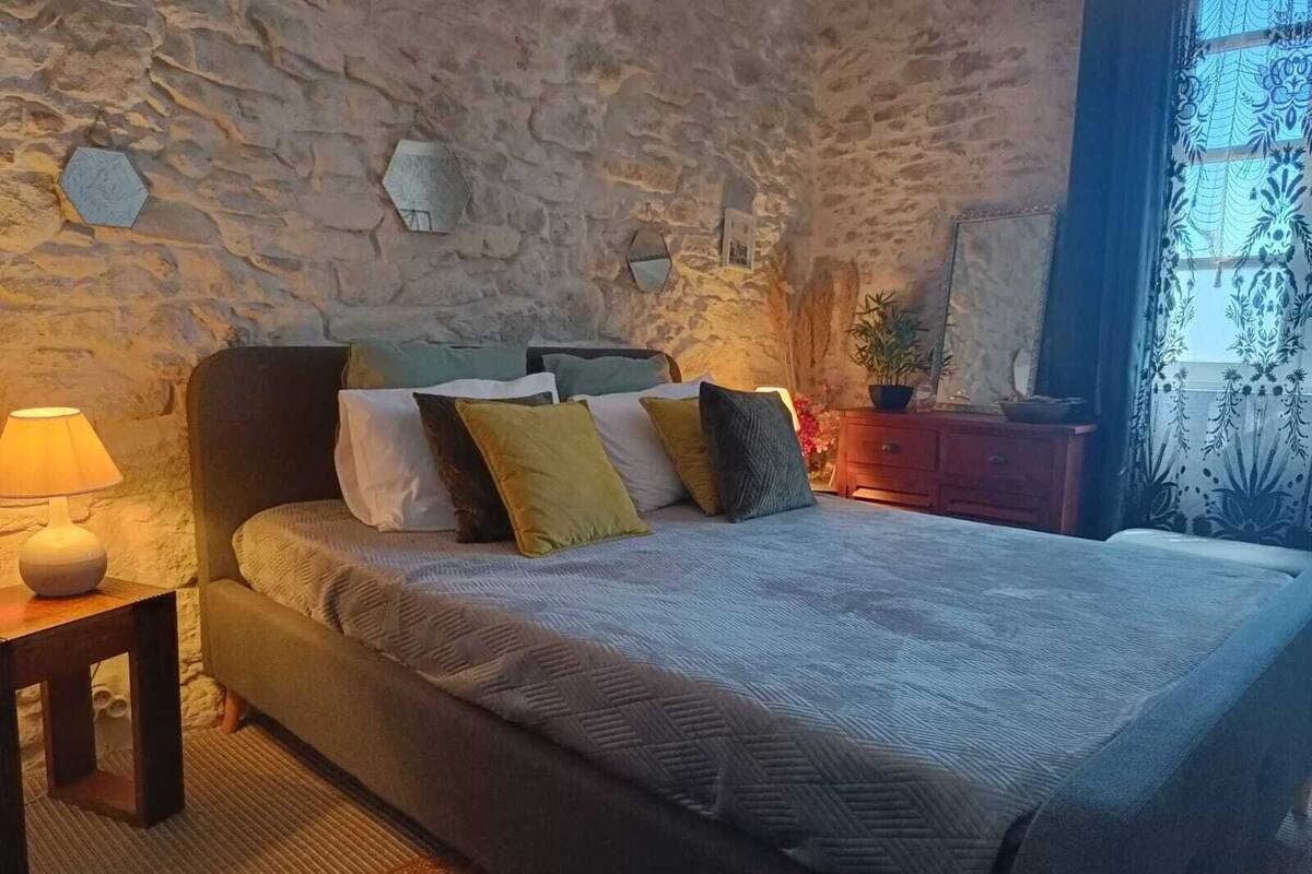 Quarto com parede de pedra, cama com várias almofadas, mesa de cabeceira com abajur e janela com cortinas.
