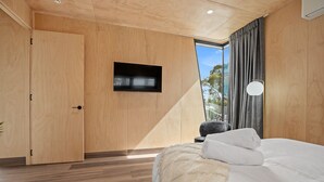 Quarto moderno com piso de madeira, uma cama grande com roupa de cama branca, uma TV fixada na parede e uma janela com cortinas.