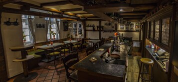 Um restaurante acolhedor com vigas de madeira, várias mesas e cadeiras, um espaço de bar e piso ladrilhado.