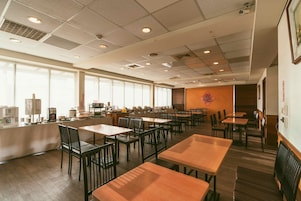 Uma sala de jantar espaçosa com várias mesas e cadeiras, janelas amplas que proporcionam luz natural, e uma área de cozinha visível ao fundo.