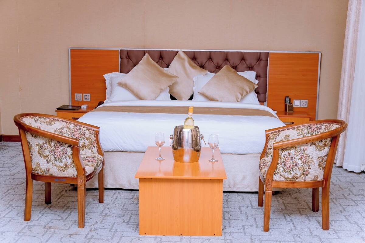 Quarto de hotel com uma cama grande, duas poltronas estofadas, uma pequena mesa de madeira e uma garrafa de champanhe.