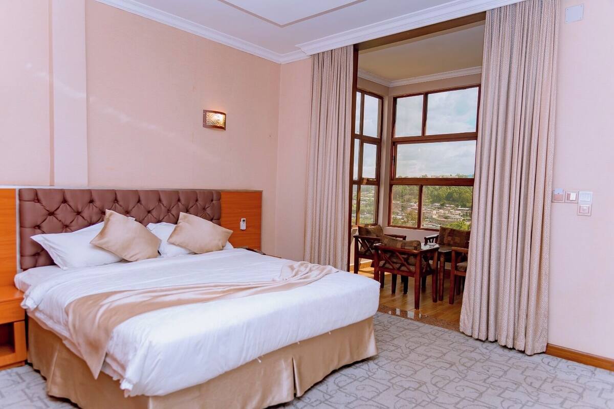 Quarto de hotel com uma cama grande, cabeceira, uma pequena mesa com cadeiras e vista pela janela.
