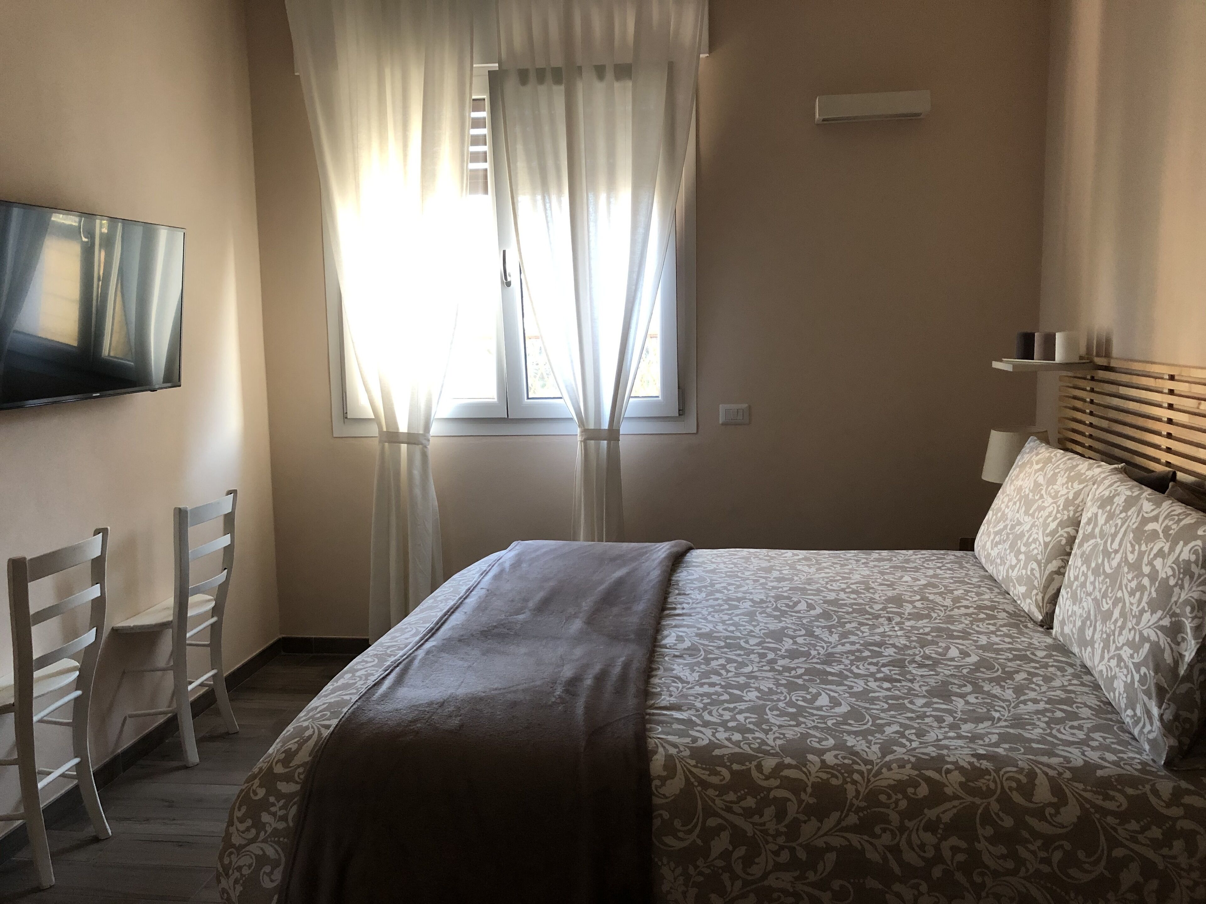 Quarto com cama, televisão na parede, janela com cortinas e mesa de cabeceira com abajur.