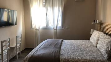 Quarto com cama, televisão na parede, janela com cortinas e mesa de cabeceira com abajur.