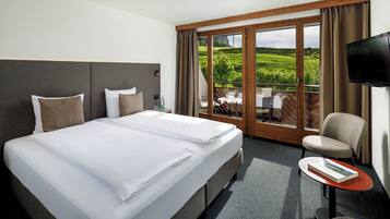 Quarto de hotel com uma cama grande, televisão, uma pequena mesa com um livro e varanda com vista para o verde.