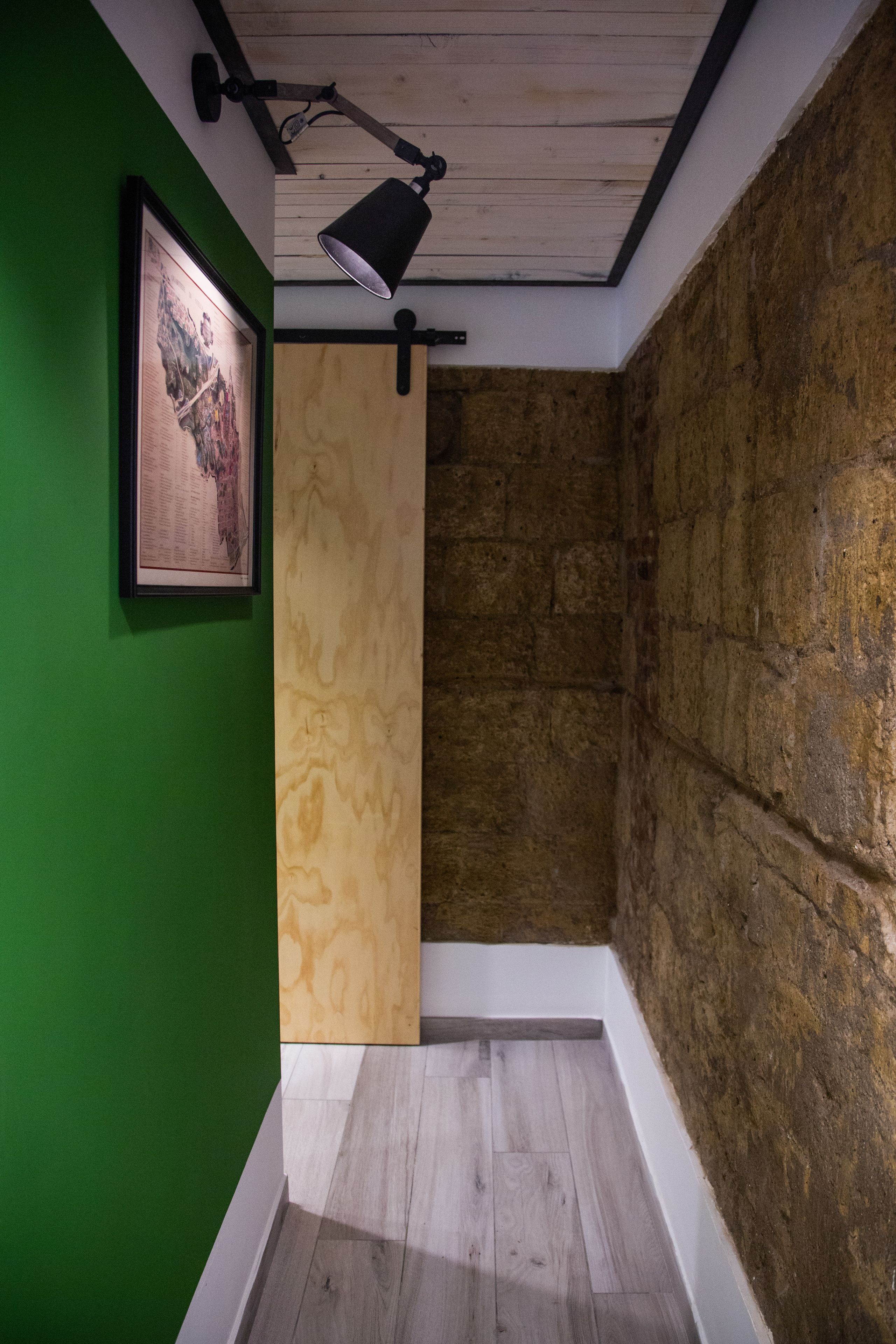 Um corredor estreito com paredes verdes, piso de madeira e um quadro emoldurado à esquerda.
