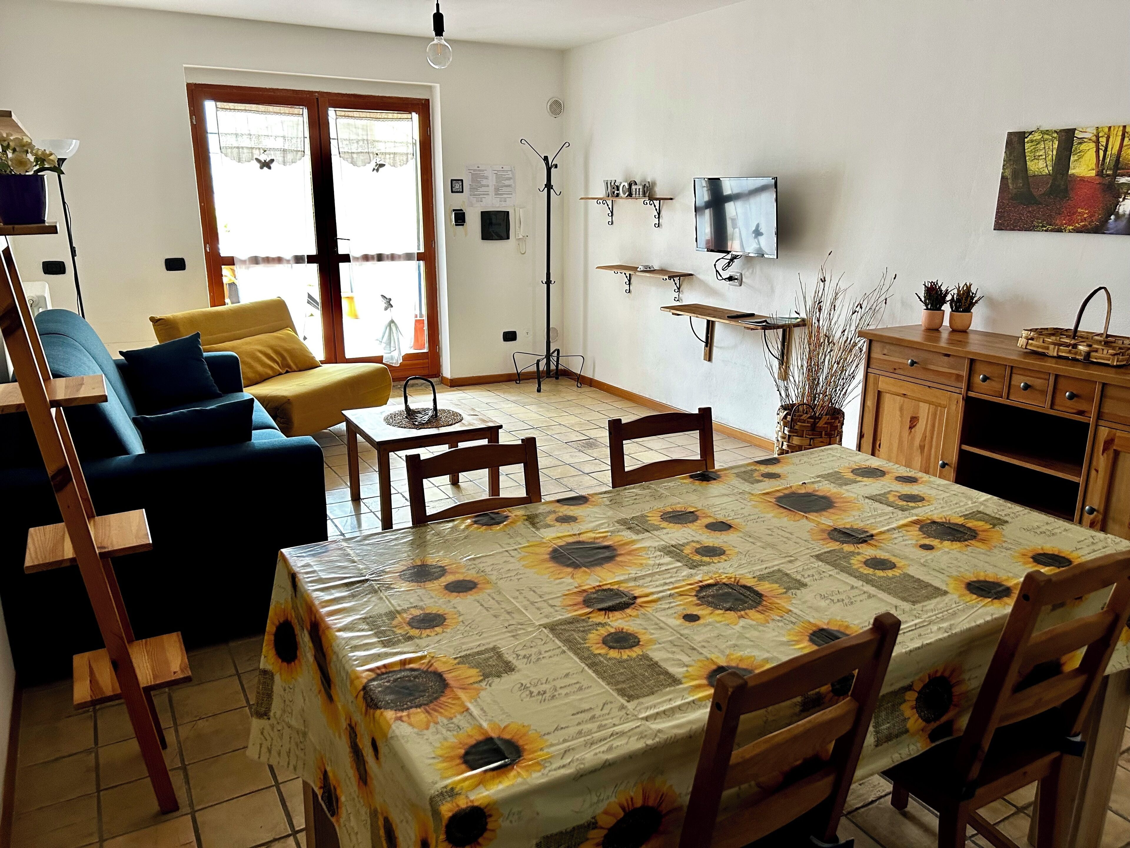 Uma sala de estar com sofá, uma mesa de jantar com toalha de mesa de girassol, TV fixada na parede e um quadro de uma floresta.