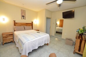 Um quarto com cama, mesas de cabeceira, ventilador de teto, televisão e espelho.
