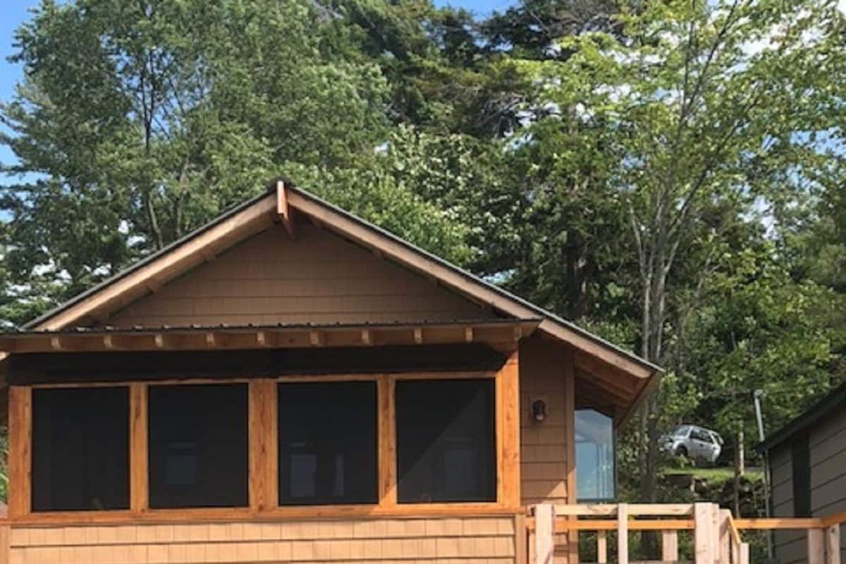 Uma cabana de madeira com um deck, churrasqueira e um veículo estacionado perto.