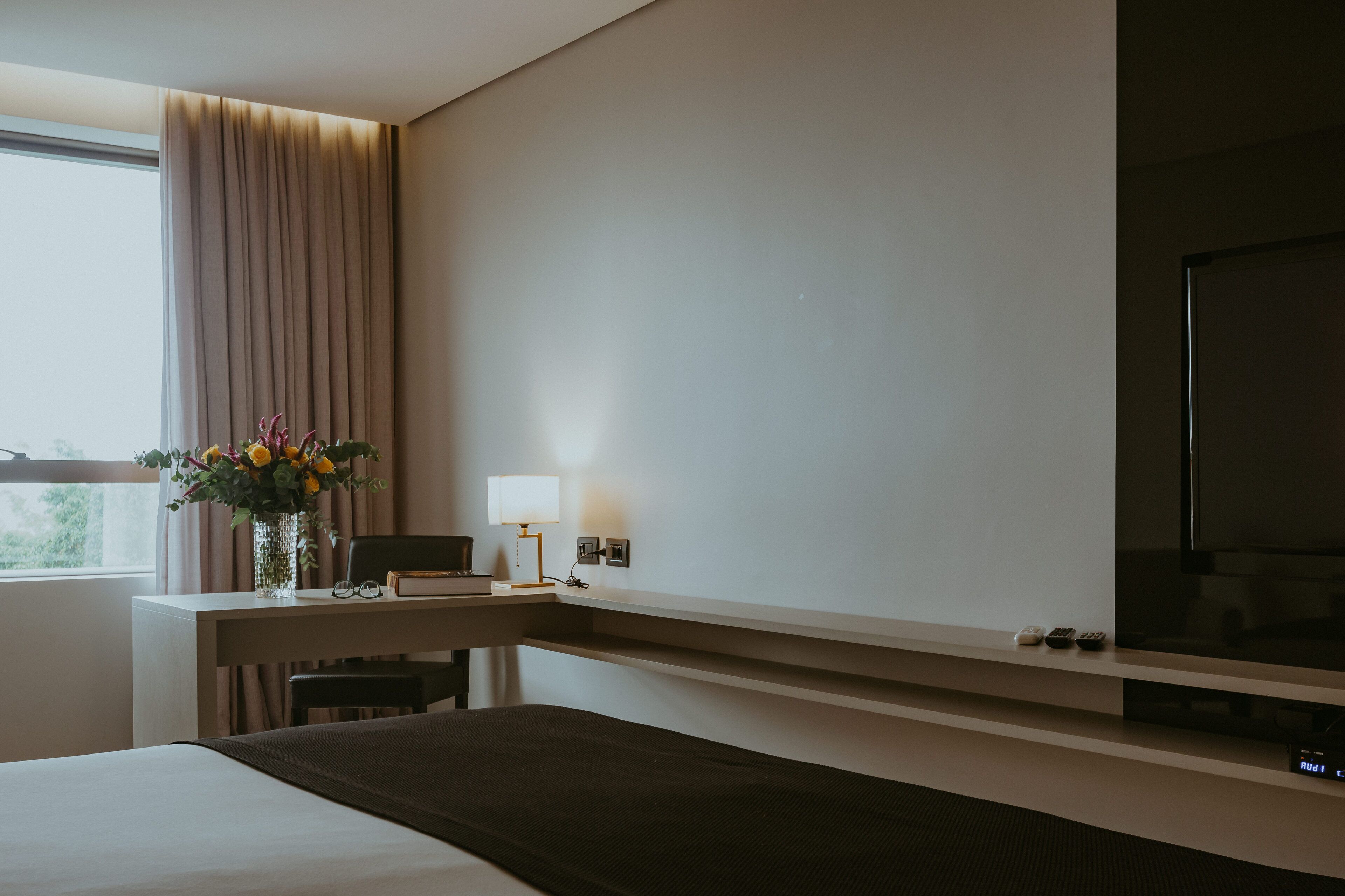 Quarto de hotel com cama, escrivaninha com luminária, televisão e janela com cortinas.