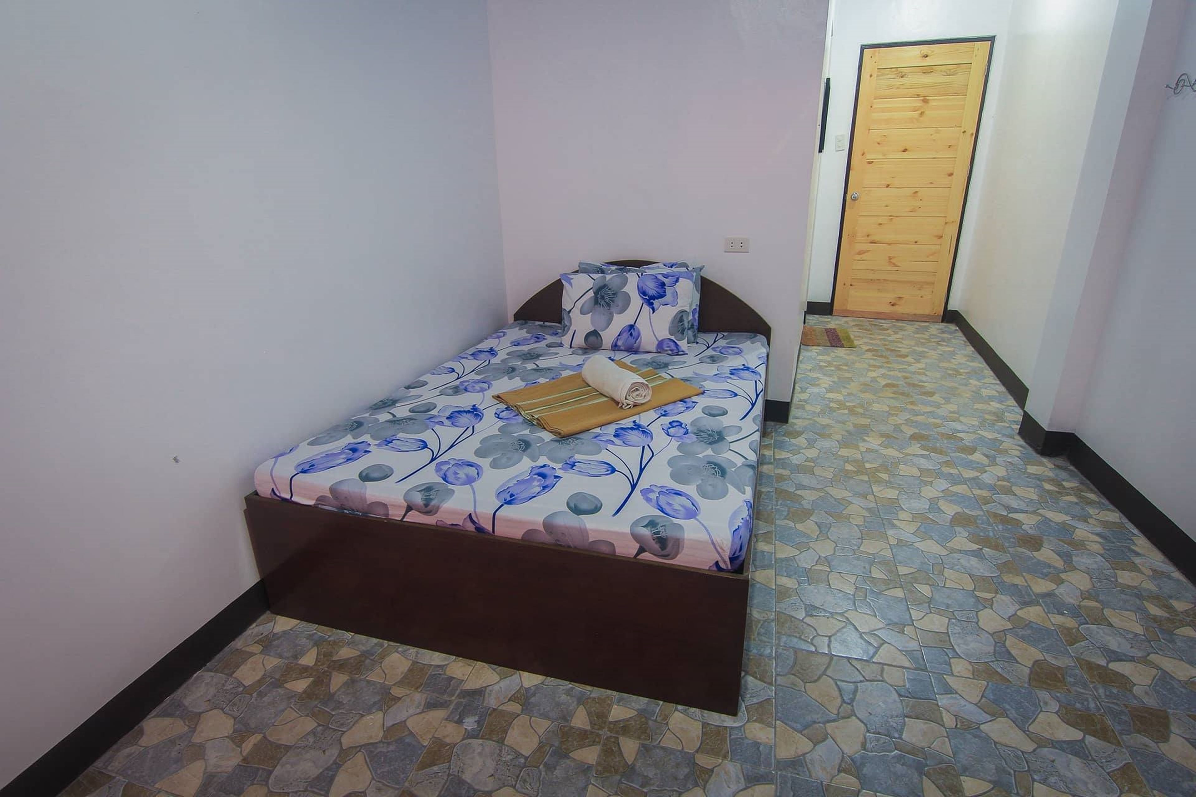 Quarto pequeno com cama, porta de madeira e ladrilhos estampados.