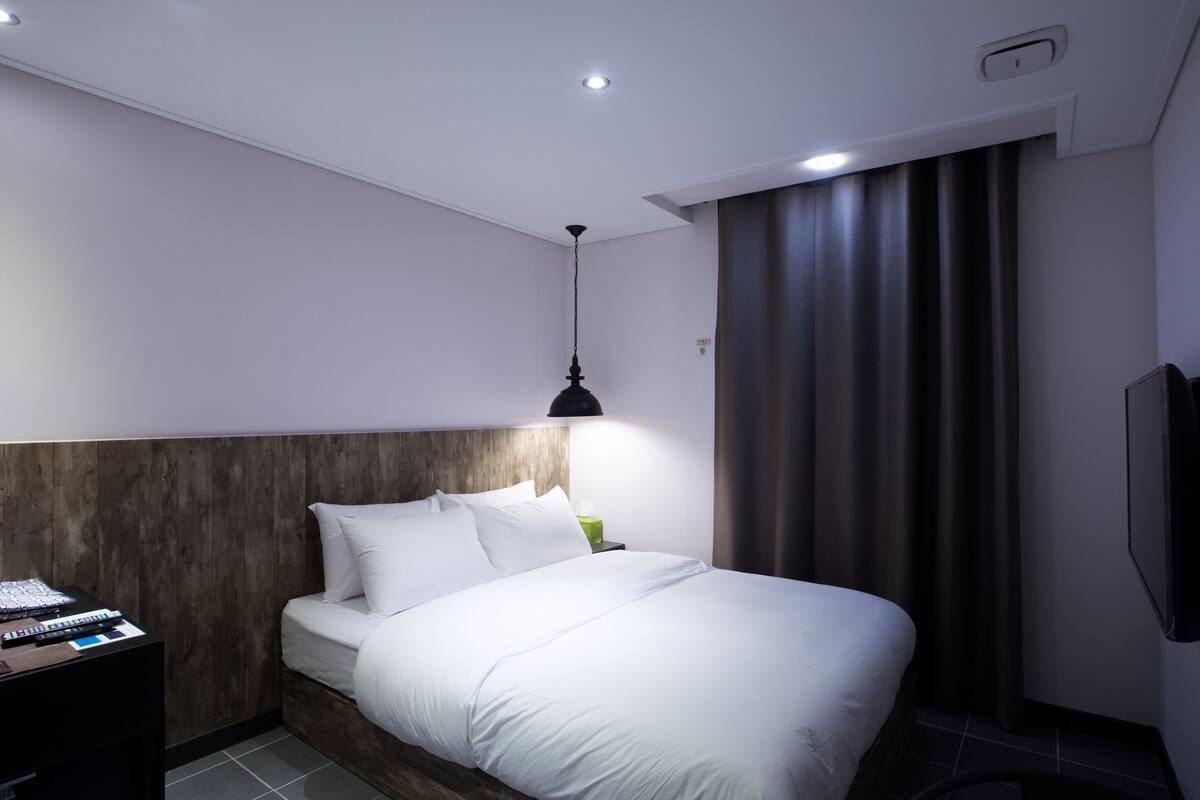 Quarto de hotel com cama, mesa de cabeceira, abajur e cortina.