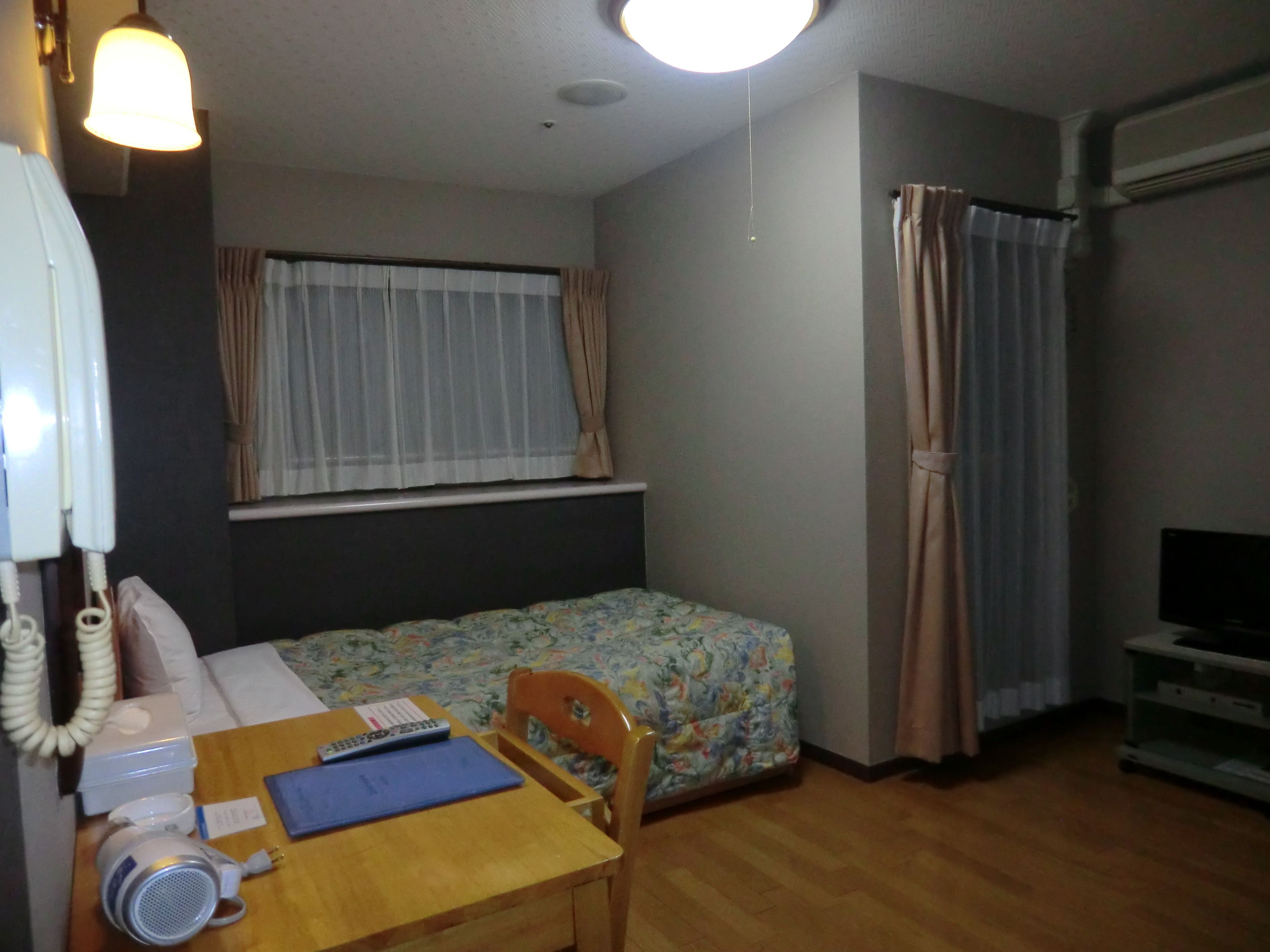 Quarto pequeno e compacto com uma cama de solteiro, uma escrivaninha com cadeira, um telefone e uma televisão.