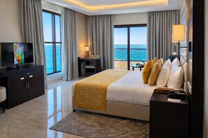 Quarto de hotel com uma cama grande, uma escrivaninha, uma televisão e vista para o oceano.