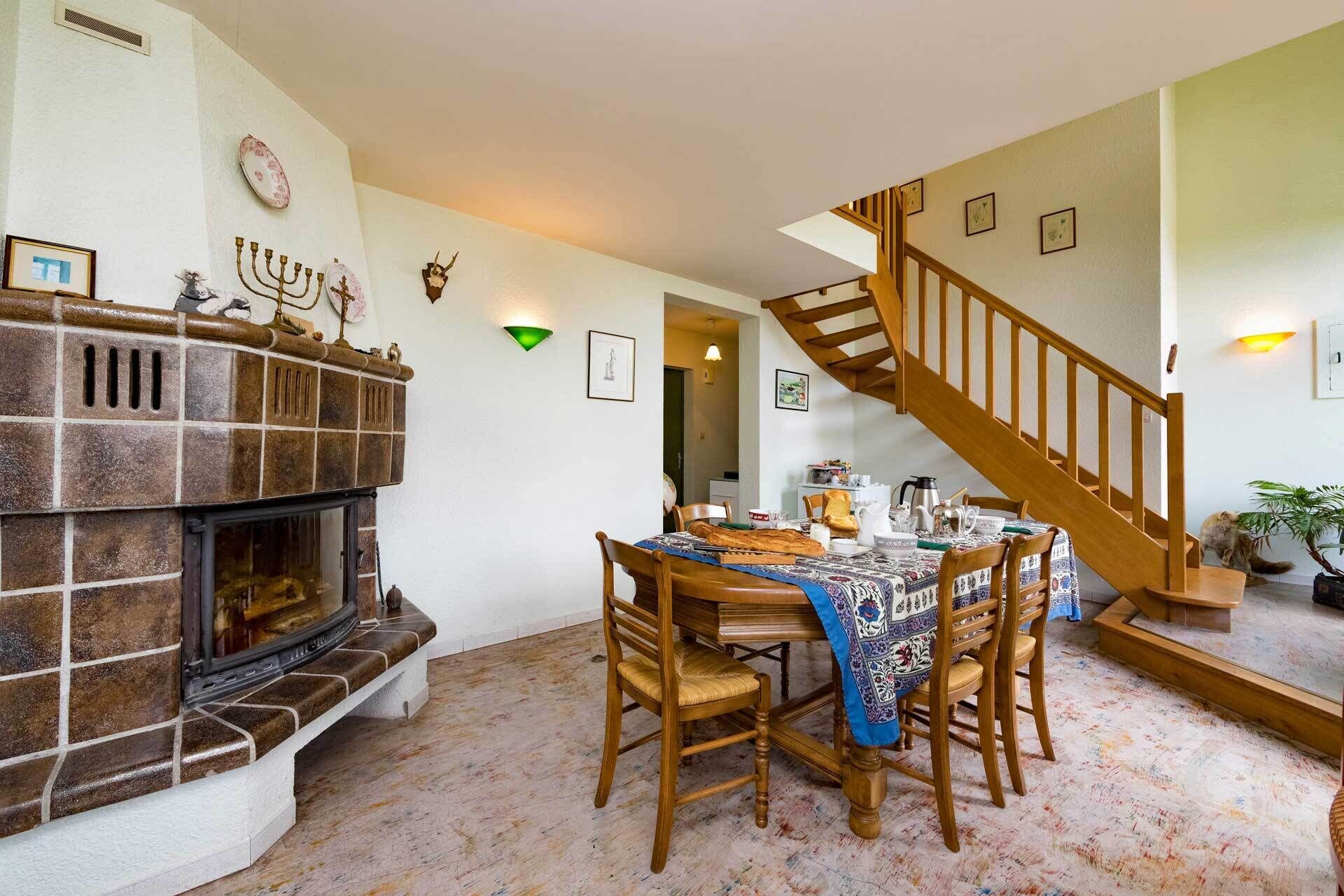 Uma sala de jantar com uma mesa de madeira, cadeiras e toalha de mesa estampada. Uma lareira com revestimento de azulejos está à esquerda. Uma escada com corrimão de madeira leva a um andar superior.