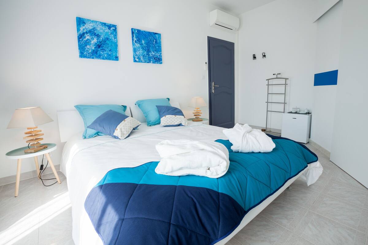 Quarto moderno com uma cama grande, duas obras de arte azuis na parede, uma porta, uma pequena mesa de cabeceira com abajur e um guarda-roupa branco.