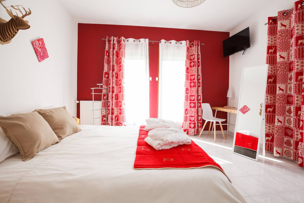 Quarto com cama, cortinas vermelhas, escrivaninha e televisão.
